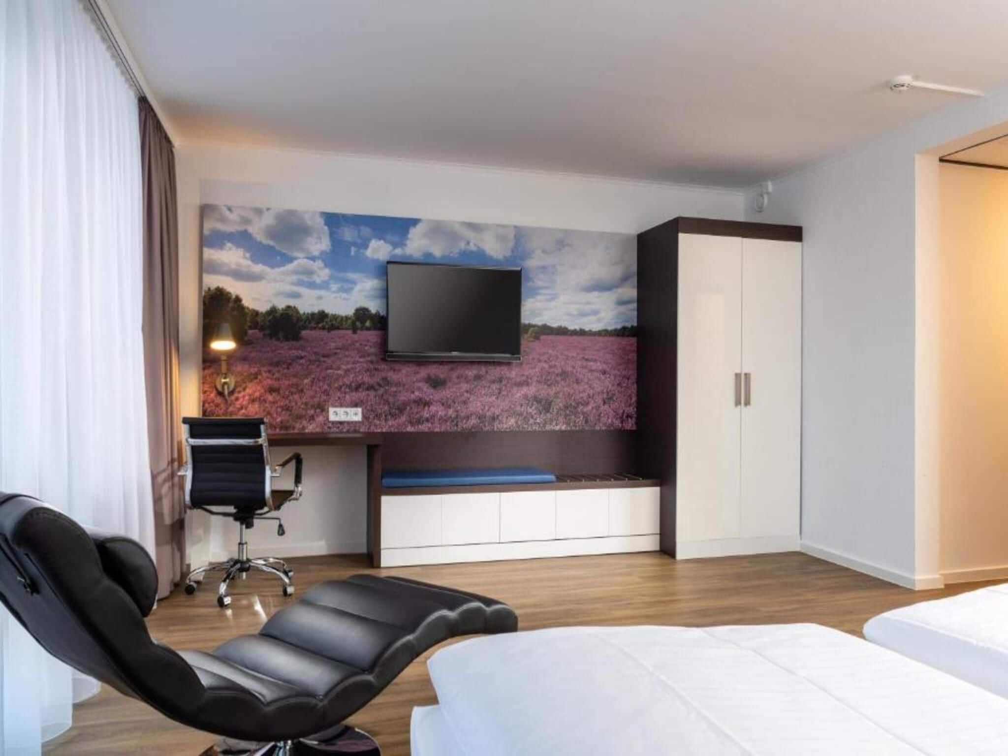 Quarto de hotel com cama, escrivaninha com cadeira, televisão na parede e guarda-roupa.