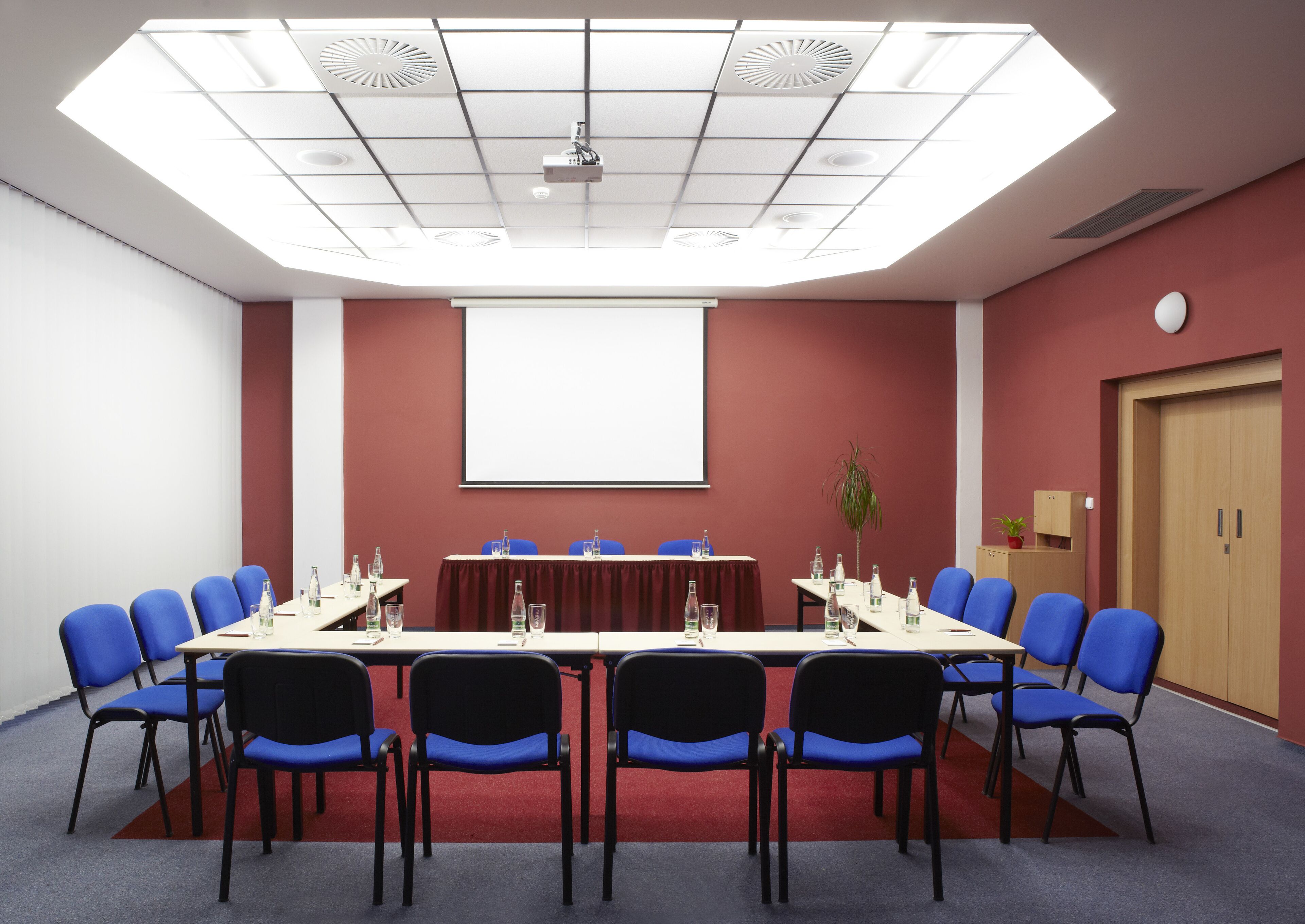 Uma sala de conferências com uma mesa grande, cadeiras azuis, uma tela de projetor e um tapete vermelho.