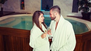Um casal em roupões relaxando numa banheira de hidromassagem, com um deck de madeira e plantas em vasos.