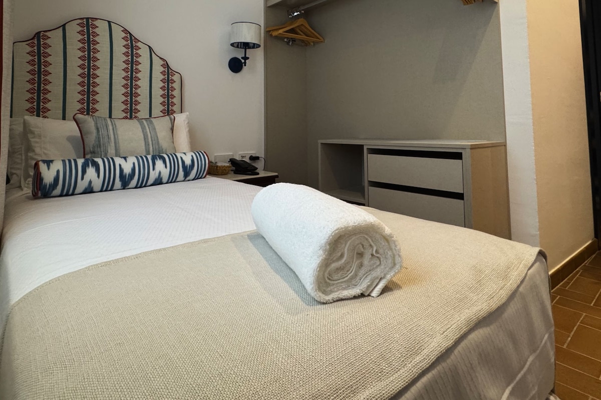 Uma cama bem arrumada com uma toalha enrolada, uma mesa de cabeceira com abajur e um guarda-roupa.