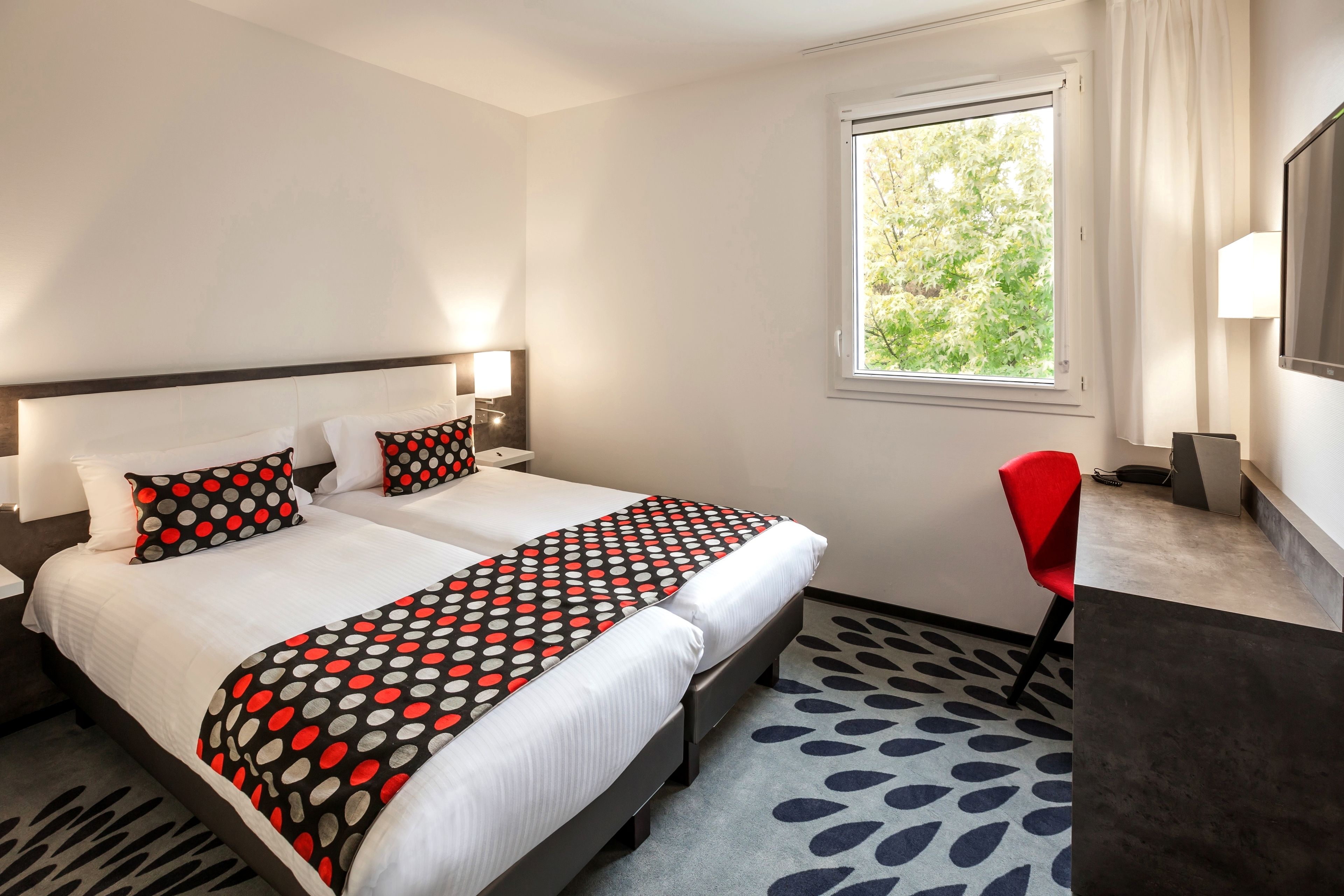 Quarto de hotel com duas camas, uma escrivaninha, uma cadeira, uma janela com vista para árvores e uma TV na parede.