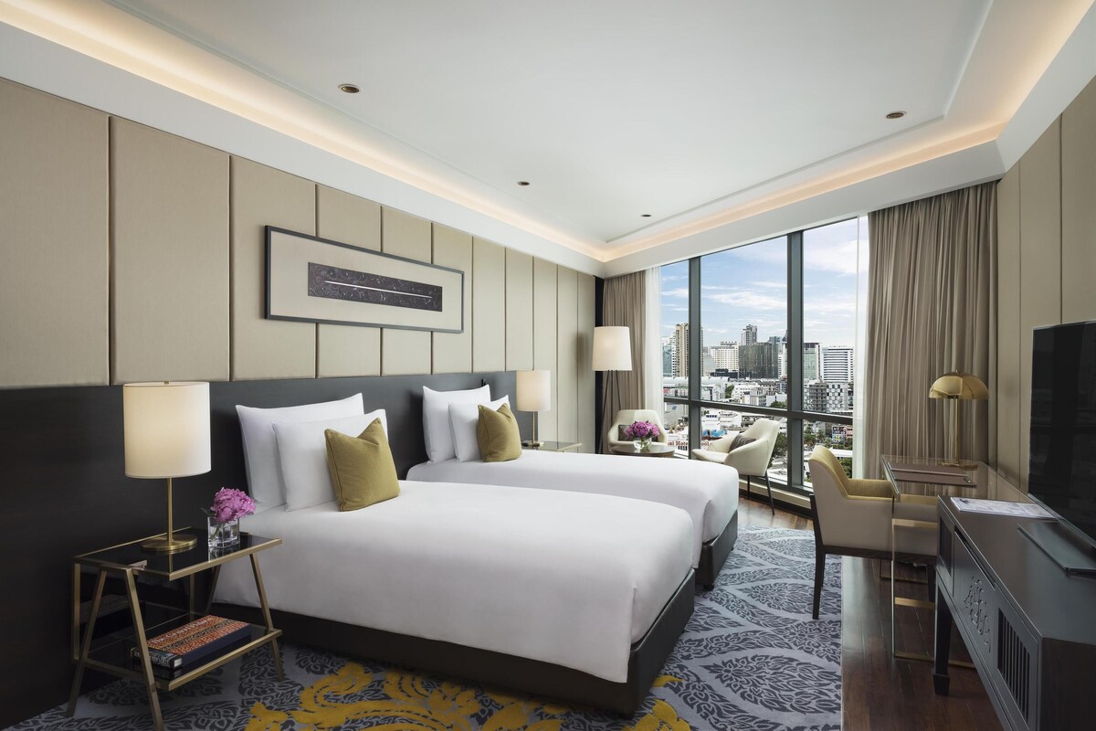 Quarto de hotel com duas camas, televisão, escrivaninha e janela grande com vista para a cidade.