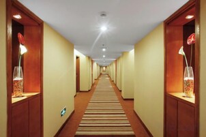 Corredor de hotel com carpete listrado, painéis de madeira e luminárias fixadas na parede.