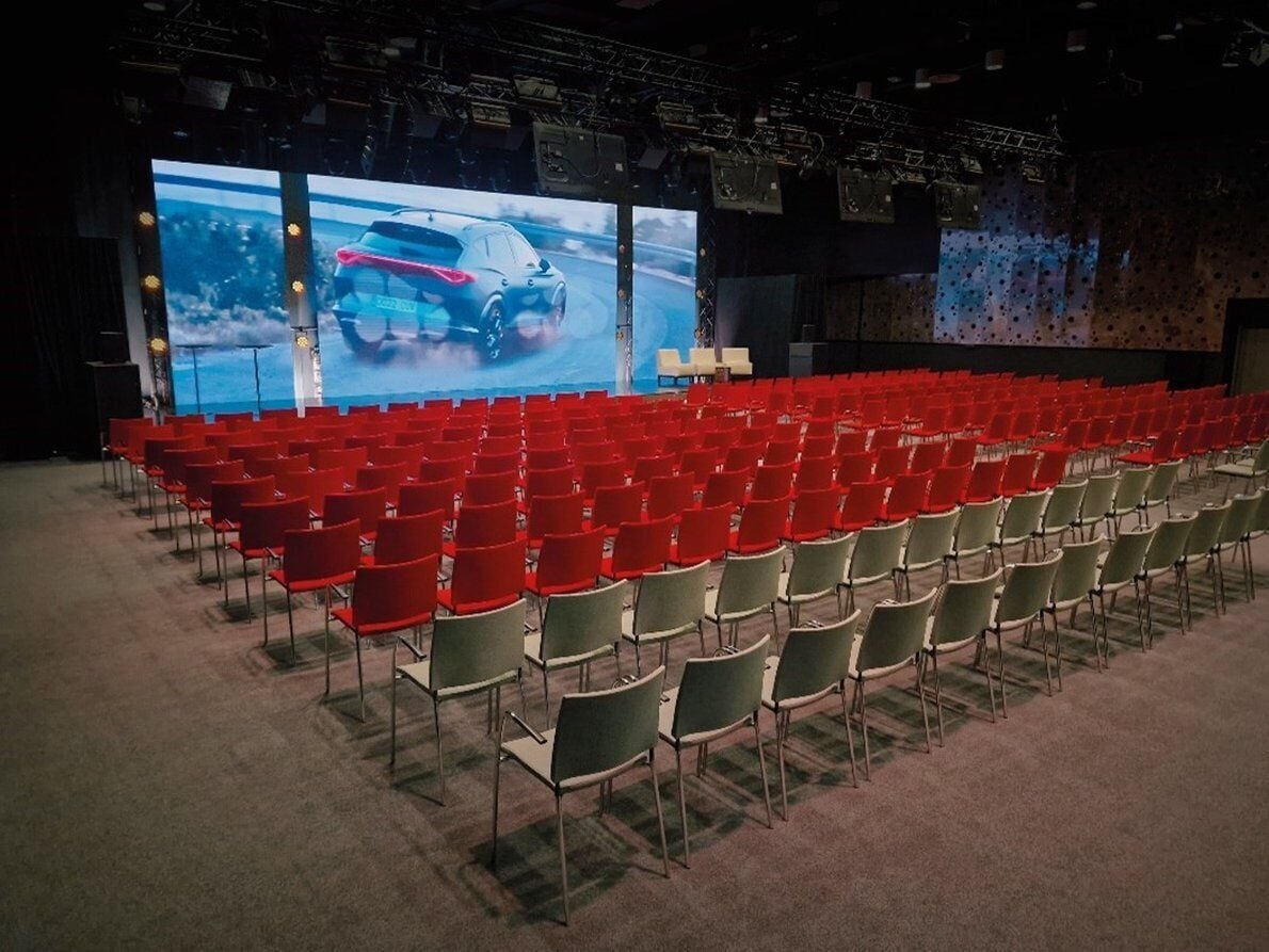 Um teatro com fileiras de cadeiras vermelhas e verdes, uma tela grande exibindo um carro e um palco com equipamentos de iluminação.