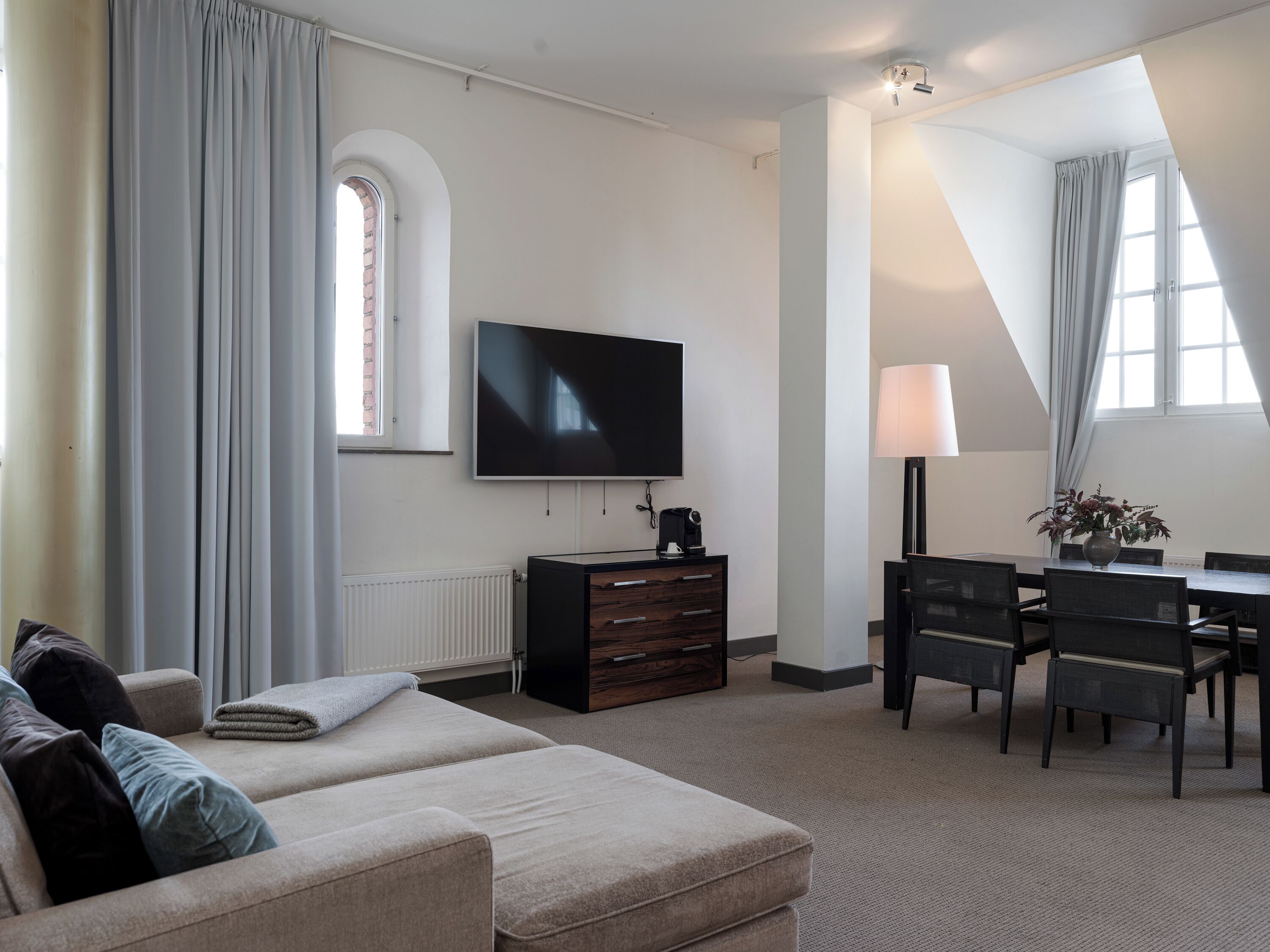 Quarto de hotel moderno com sofá, televisão de tela plana, mesa de jantar com cadeiras e luminária.