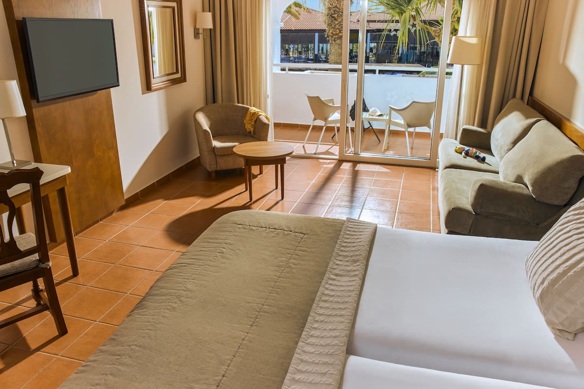 Quarto de hotel com cama, sofá, poltrona, mesa pequena, TV e vista para a piscina e palmeiras.