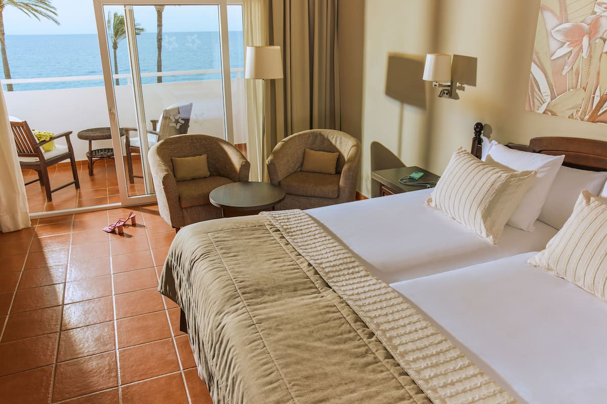 Quarto de hotel com cama, duas poltronas, uma mesinha e vista para o oceano.
