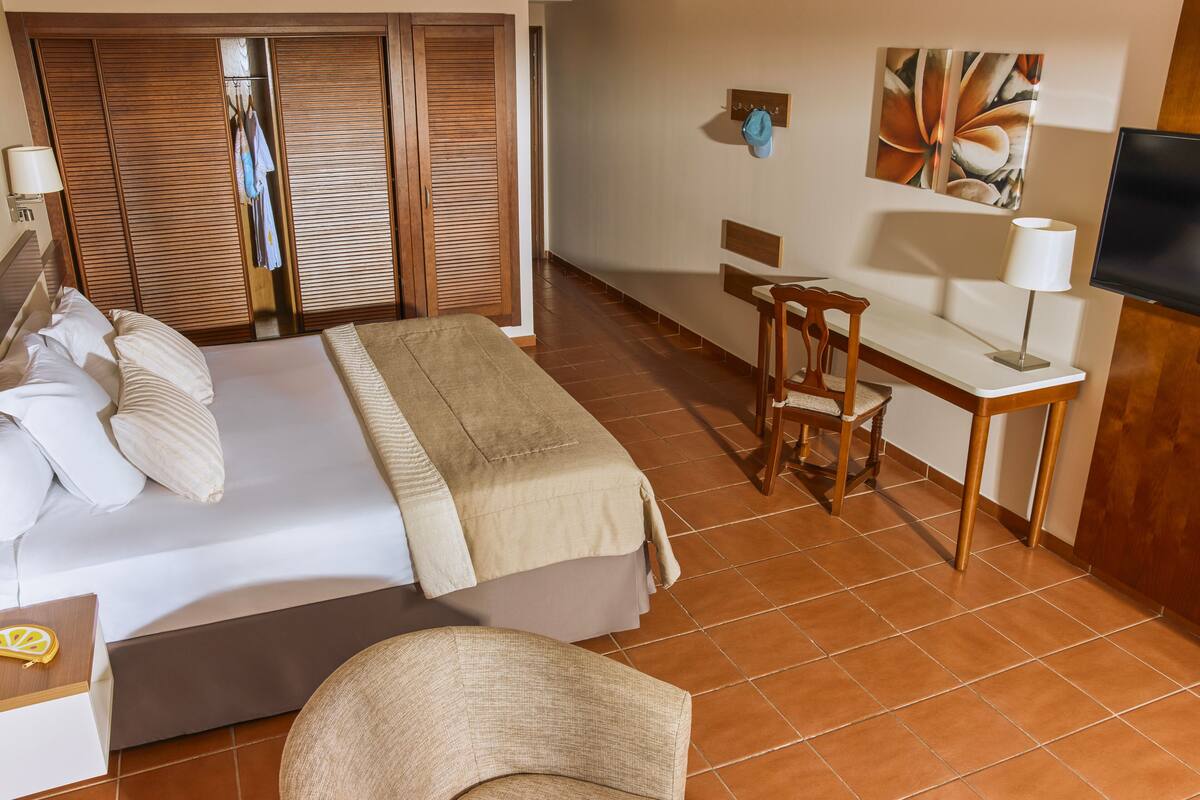 Quarto de hotel com cama, escrivaninha, cadeira, televisão e guarda-roupa.