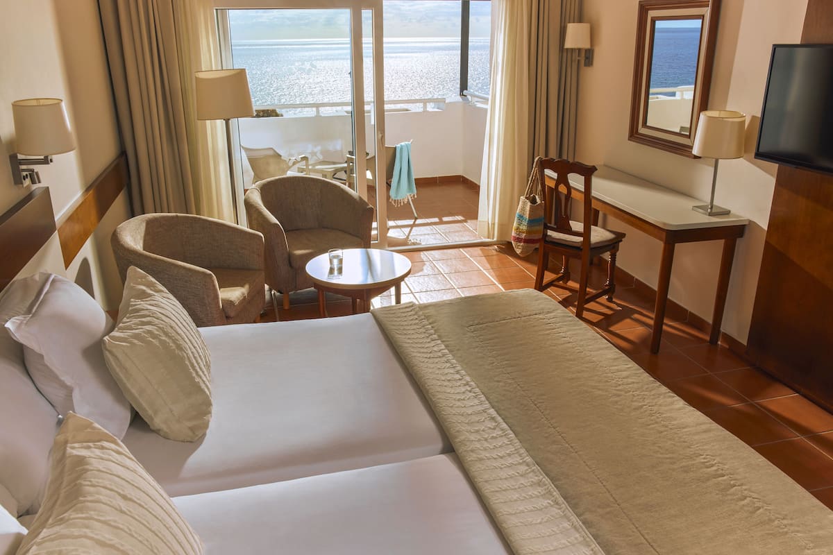 Quarto de hotel com uma cama, duas cadeiras, uma mesinha, uma escrivaninha, uma televisão e vista para o mar.