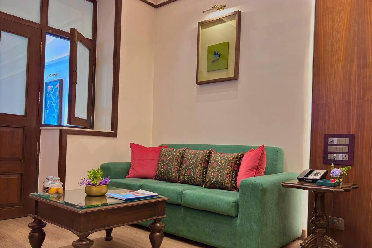 Um sofá verde com almofadas vermelhas e florais, uma mesa de centro de madeira com um livro e um vaso, e um quadro na parede.