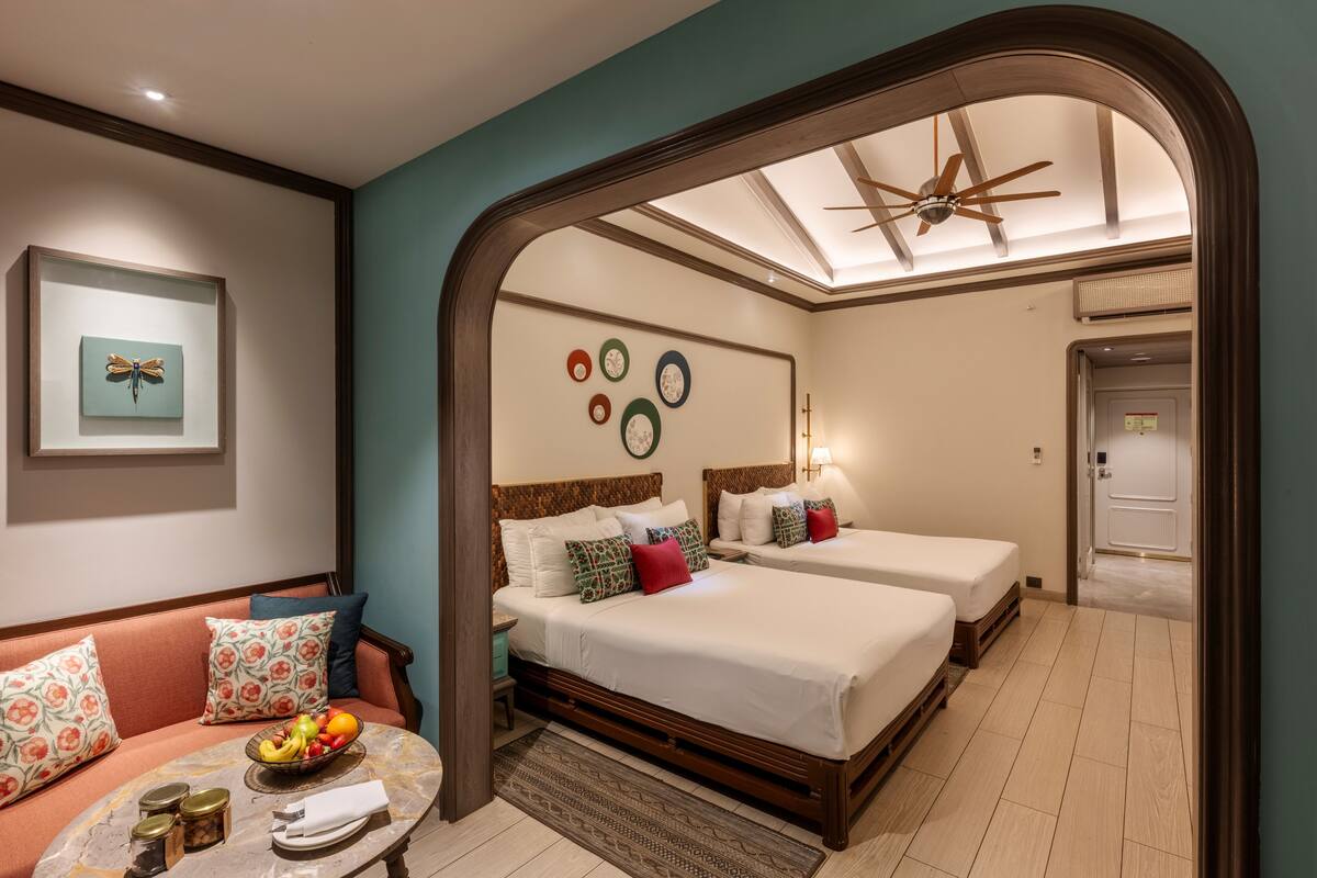 Quarto de hotel com duas camas, um sofá, uma mesa de jantar com frutas e acompanhamentos, e um ventilador de teto.