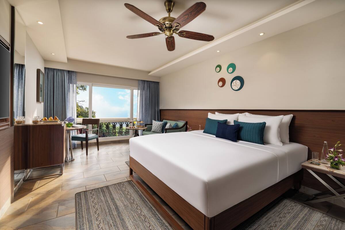 Um quarto de hotel espaçoso com uma cama grande, um ventilador de teto, uma varanda com vista e uma pequena área de estar com mesa e cadeiras.