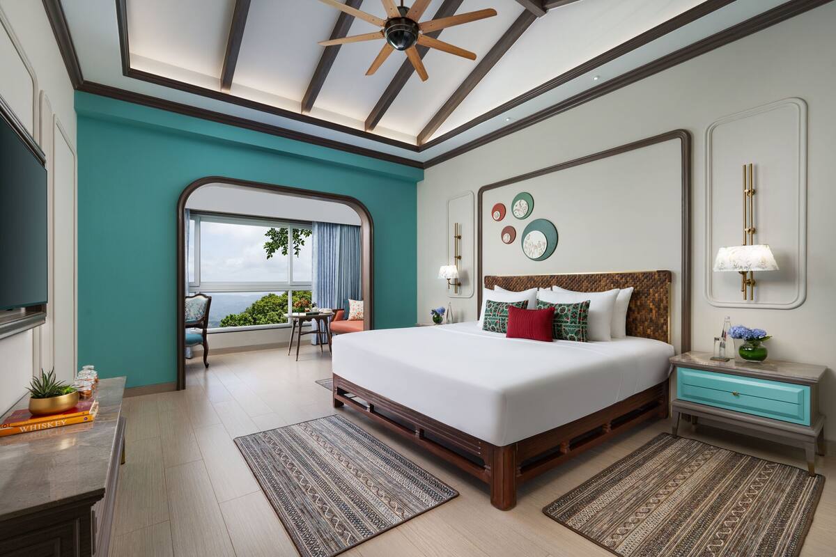 Quarto moderno com cama grande, escrivaninha com planta, vista para o exterior e ventilador de teto.