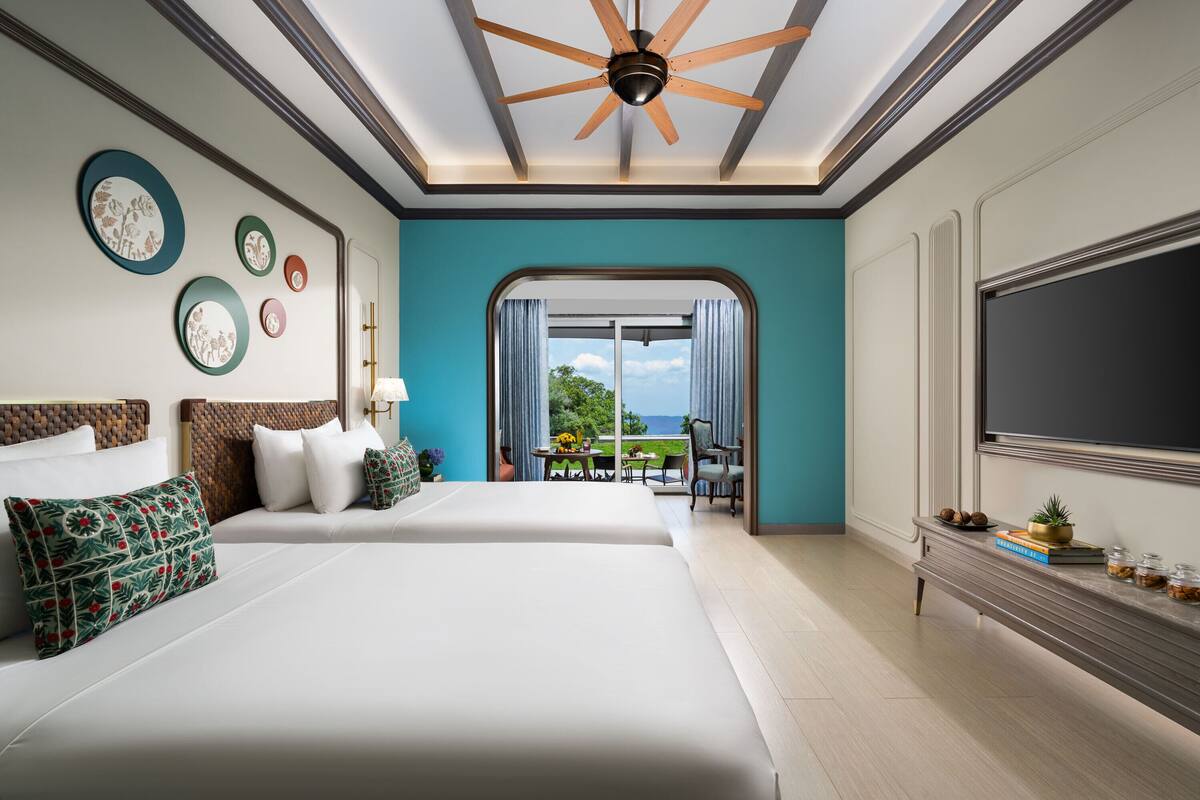 Quarto de hotel moderno com duas camas, ventoinha no teto, televisão grande e varanda com vista.