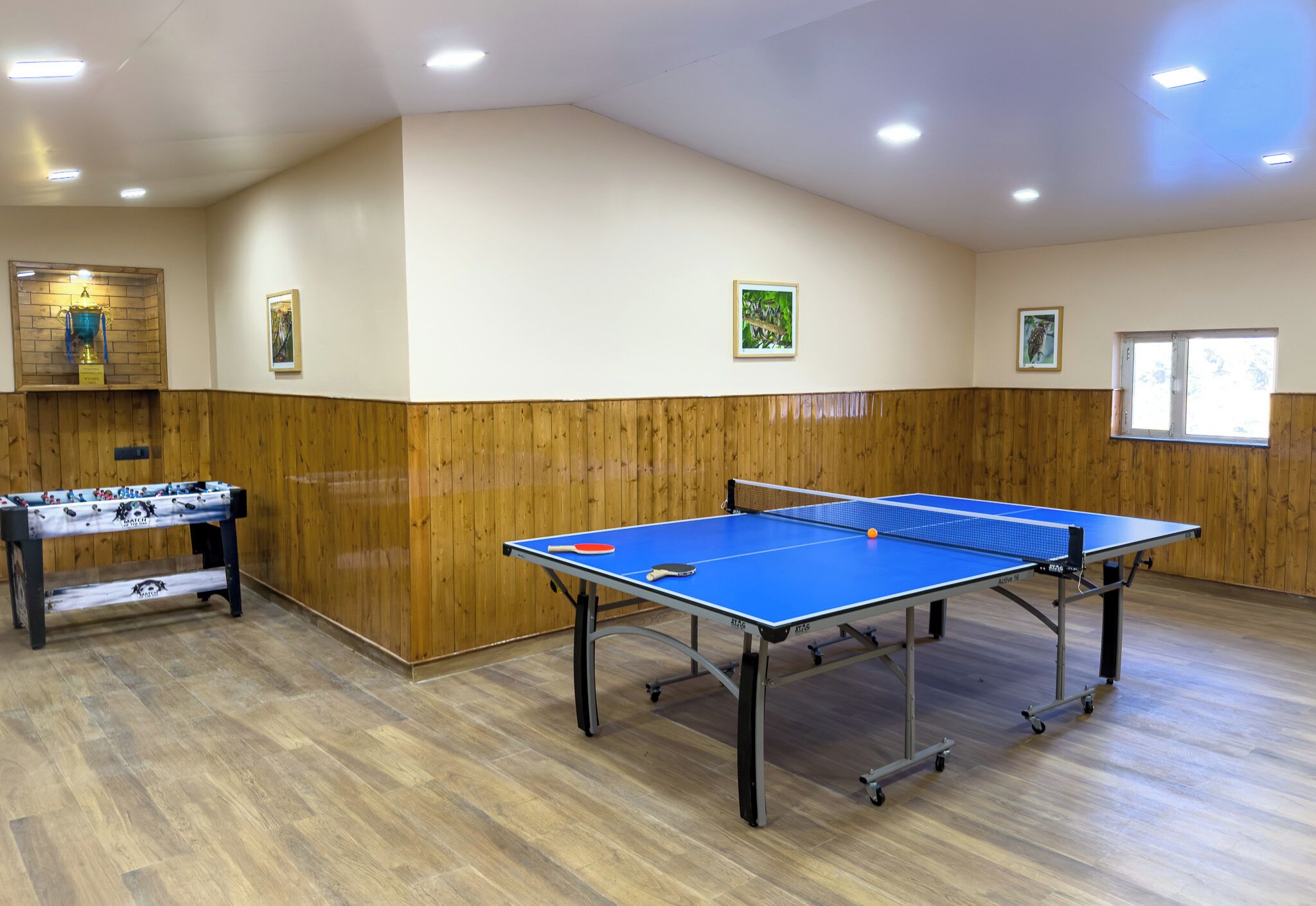 Um quarto com mesa de pingue-pongue, mesa de futebol de mesa, painéis de madeira e quadros nas paredes.