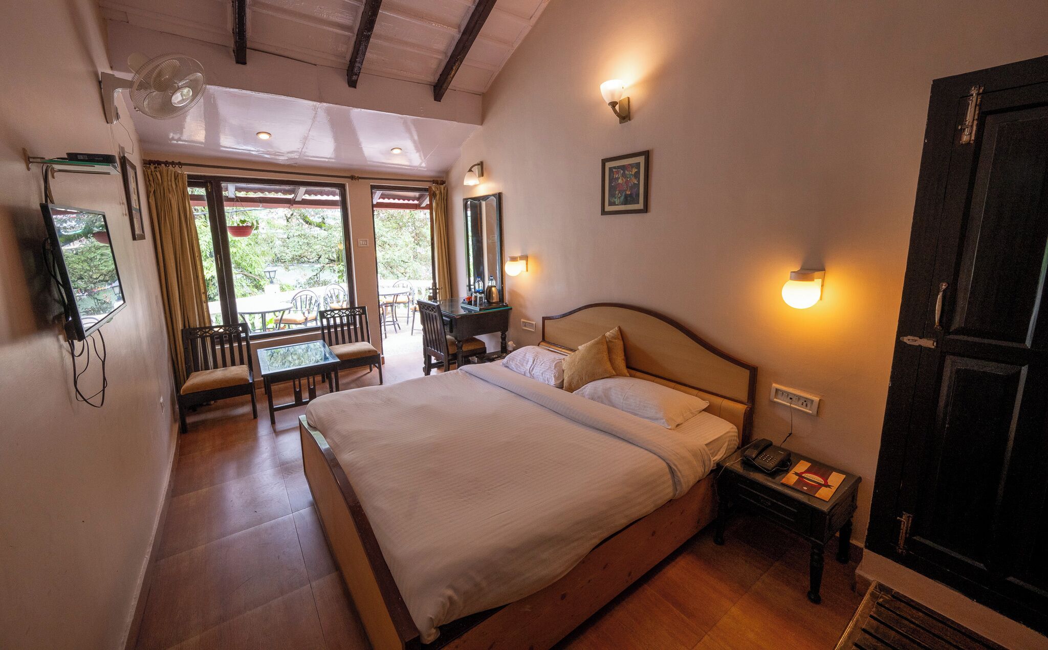 Quarto com uma cama grande, uma escrivaninha com cadeira, um ventilador e vista para o exterior.
