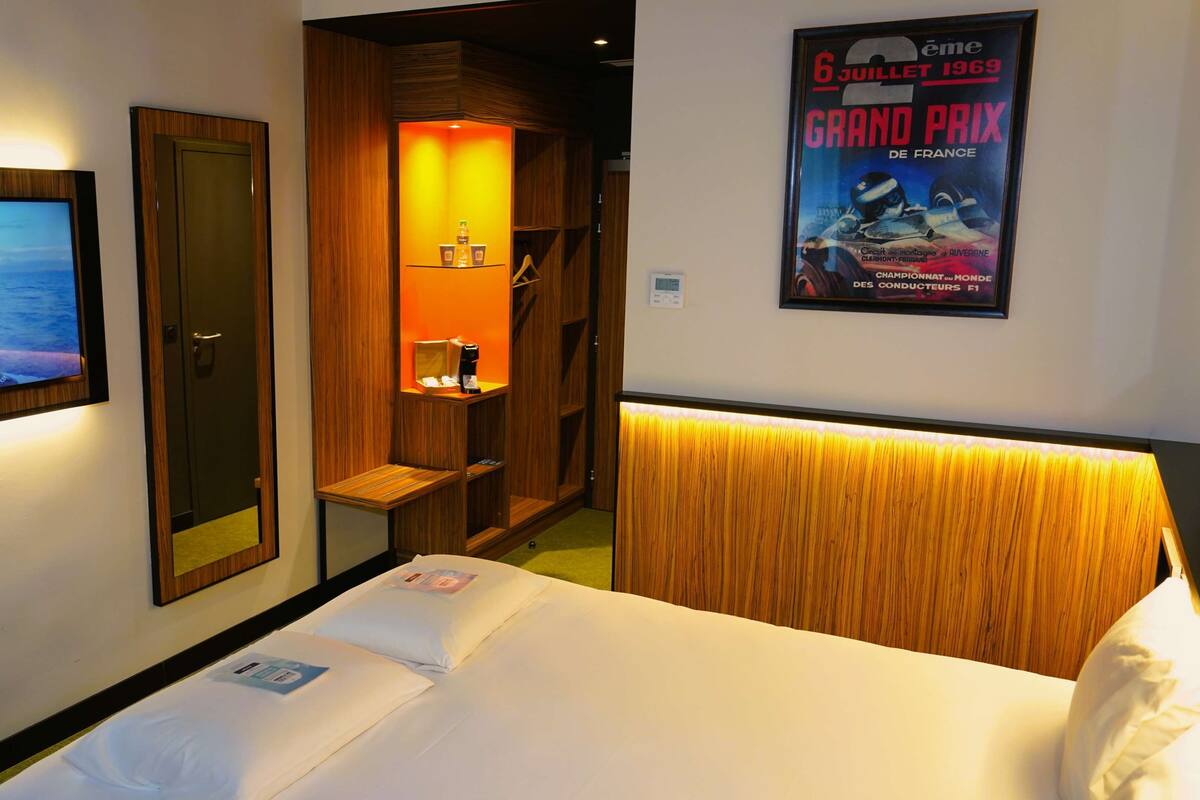Quarto de hotel com cama, televisão, guarda-roupa de madeira e um cartaz na parede.