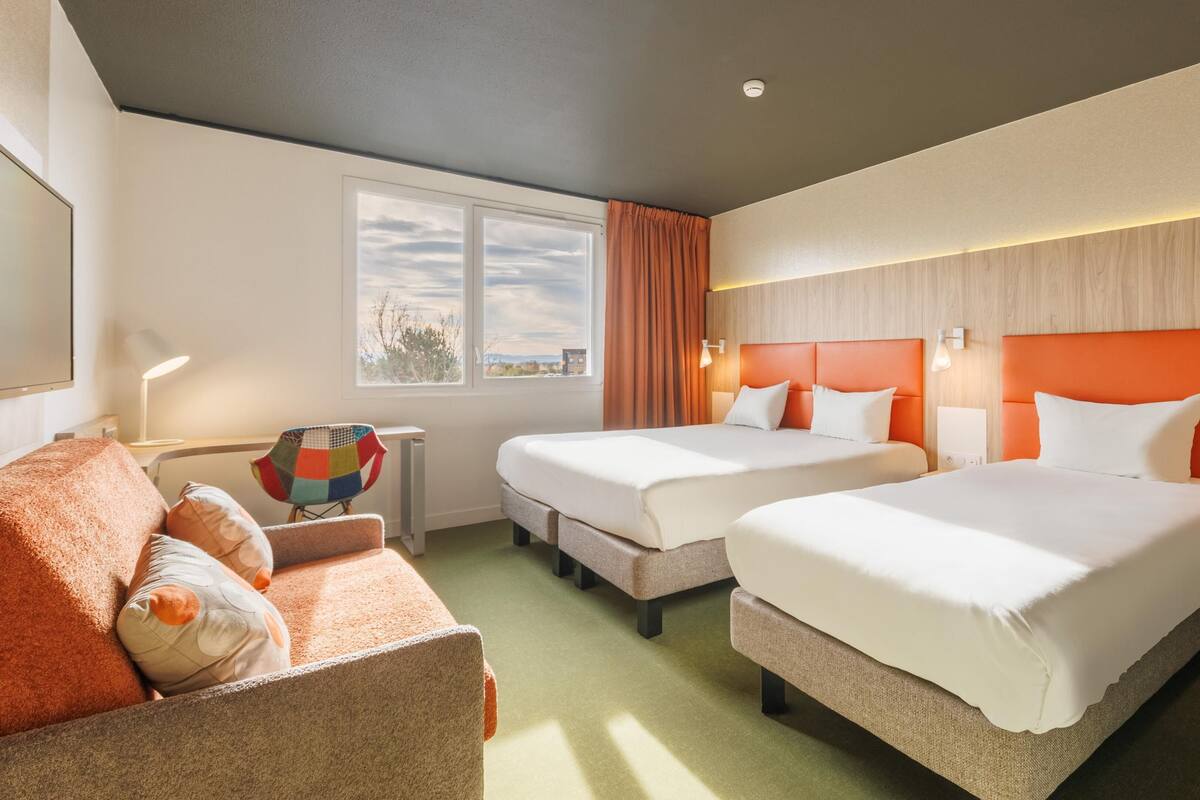 Quarto de hotel com duas camas, uma escrivaninha, uma cadeira e uma janela com vista.