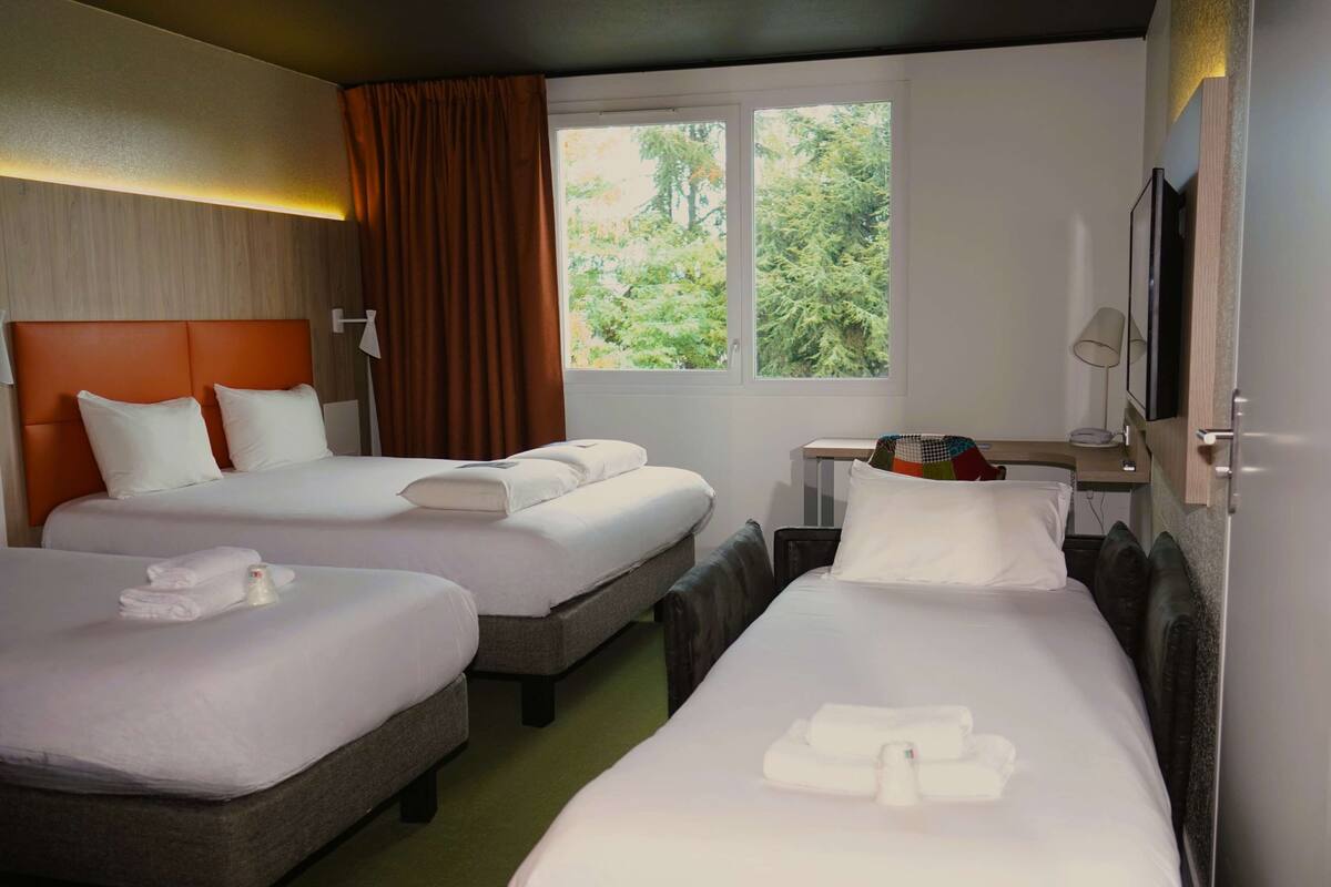 Quarto de hotel com duas camas, uma mesa, uma televisão e uma janela com vista para árvores.