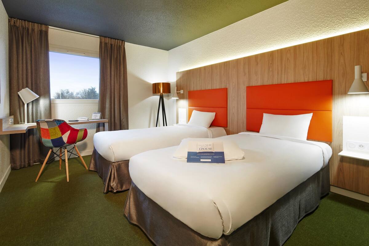 Quarto de hotel com duas camas, uma escrivaninha, uma cadeira e uma janela com cortinas.
