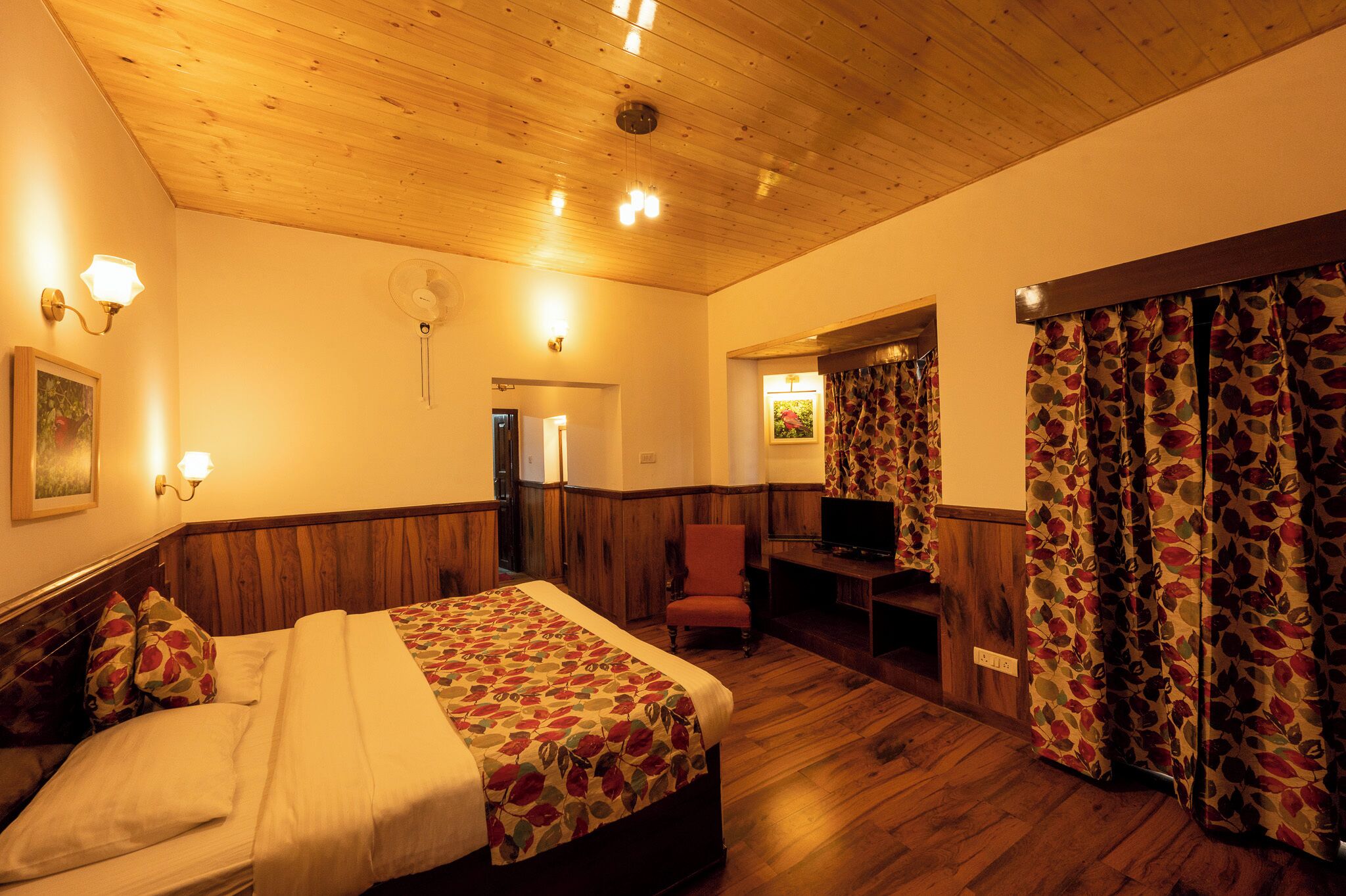 Quarto de hotel com teto de madeira, cama com roupa de cama florida, uma cadeira, escrivaninha com televisão e janela com cortinas florais.