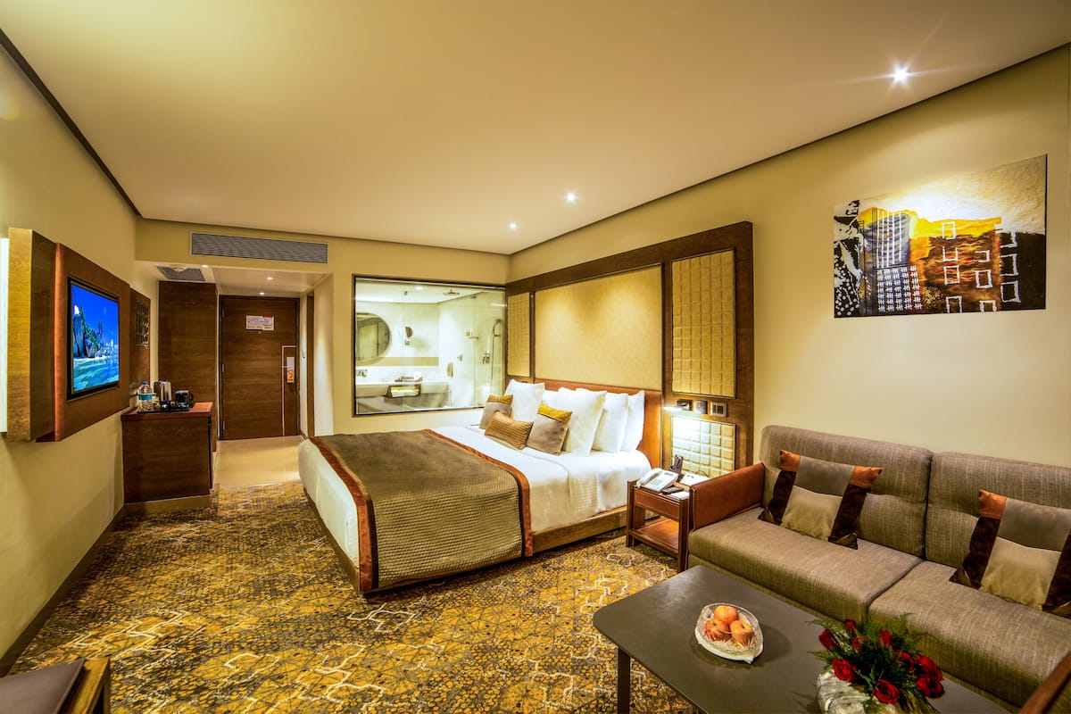 Quarto de hotel com cama, sofá, mesa de centro com frutas, televisão e banheiro visível por uma porta aberta.