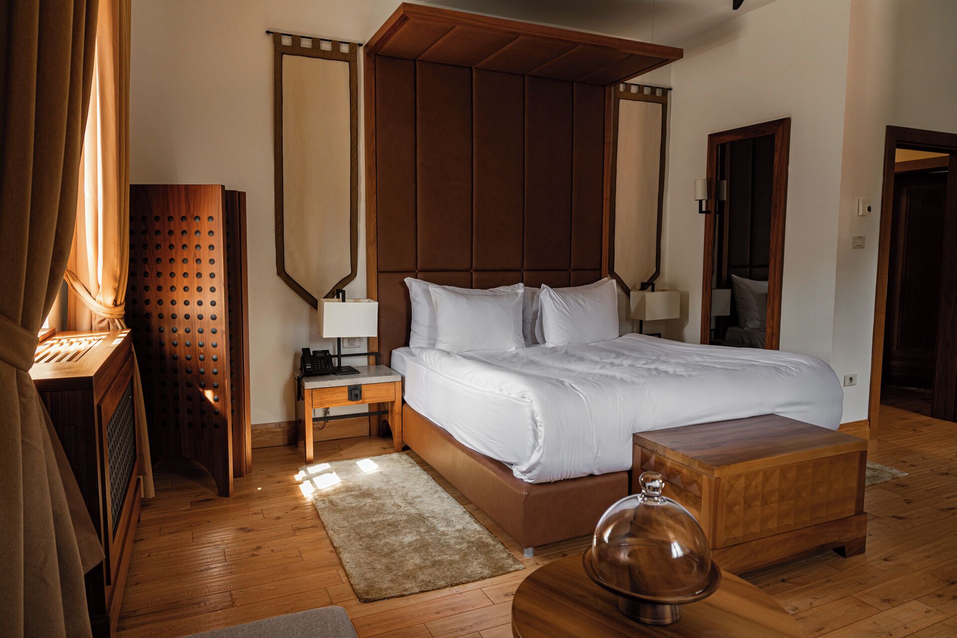Quarto de hotel com uma cama grande, cabeceira de madeira, mesas de cabeceira e um criado-mudo.