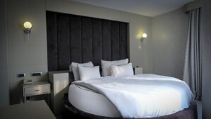 Quarto de hotel moderno com uma cama grande, duas mesas de cabeceira, um painel de cabeceira escuro e iluminação embutida na parede.