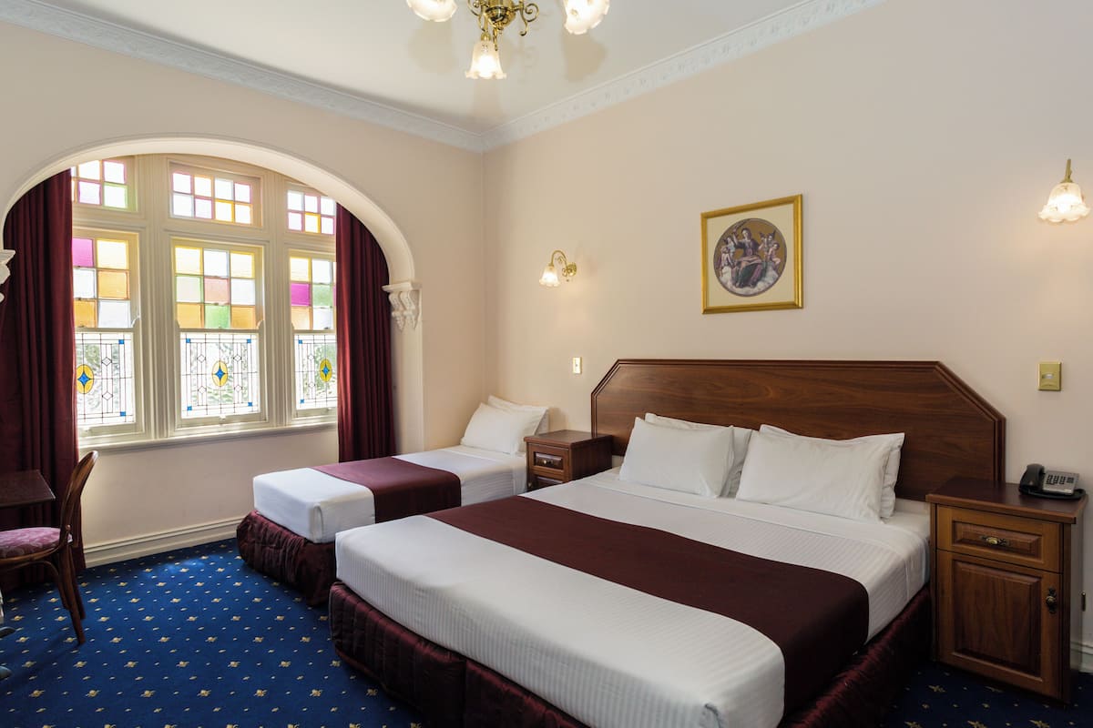 Quarto de hotel com duas camas, cabeceira de madeira, mesa de cabeceira, uma cadeira, janela com vitrais e um quadro fixado na parede.