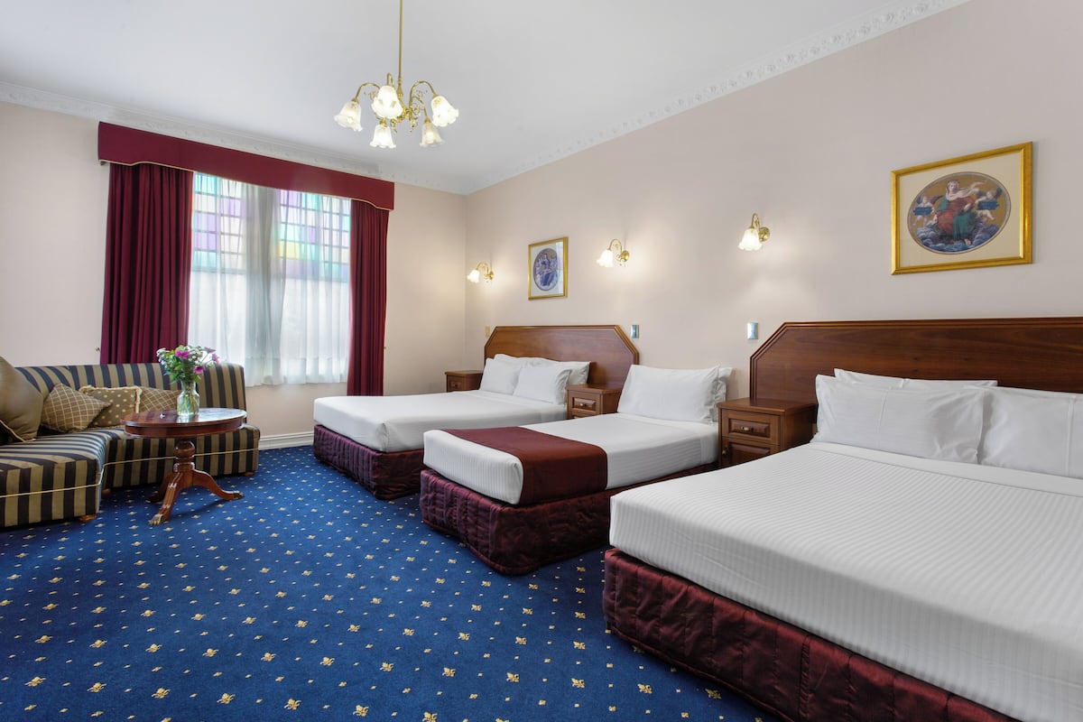 Quarto de hotel com duas camas de solteiro, um sofá, uma mesinha com um vaso e uma janela grande com cortinas.