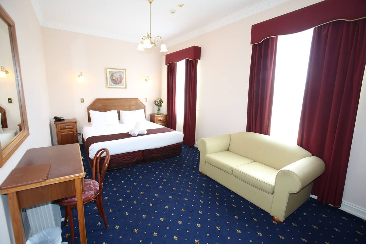 Quarto de hotel com cama, escrivaninha, poltrona, sofá, lustre e uma janela grande com cortinas.