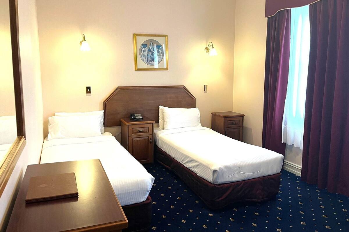 Quarto de hotel com duas camas de solteiro, uma mesinha, um criado-mudo com telefone e um quadro na parede.