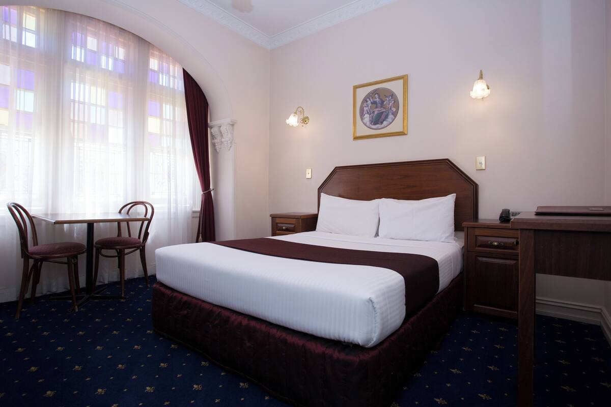 Quarto de hotel com uma cama grande, uma escrivaninha, uma cadeira e uma mesinha.