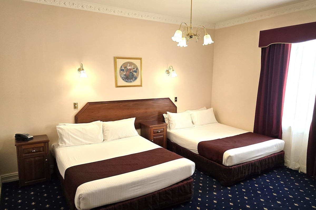 Quarto de hotel com duas camas, um candelabro, um quadro na parede e uma janela com cortinas.