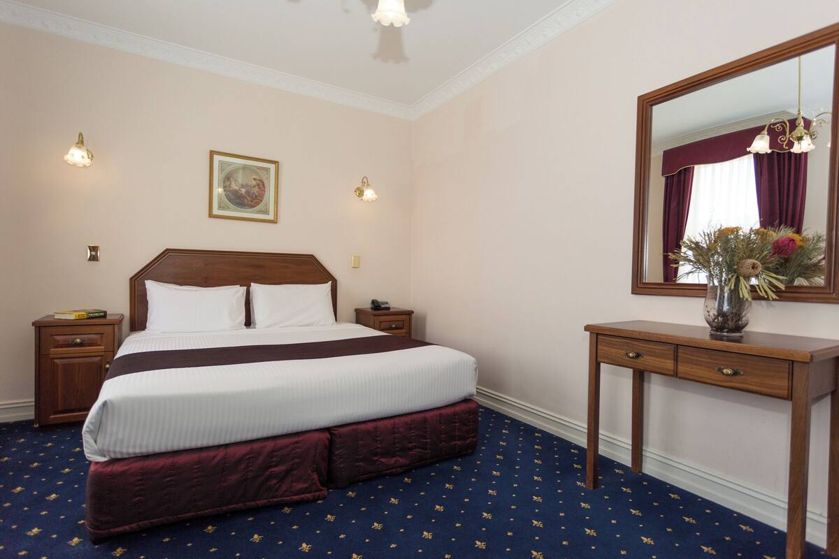 Quarto de hotel com uma cama grande, duas mesas de cabeceira, uma cômoda, um espelho e um quadro na parede.
