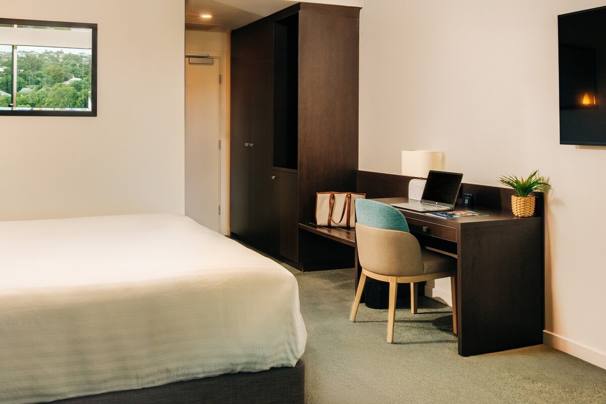 Quarto de hotel com uma cama, uma mesa com um laptop, uma cadeira, um abajur e uma pequena planta.