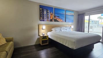 Quarto de hotel com uma cama grande, mesa de cabeceira, luminária, cadeira e janela com vista para a praia e edifícios.