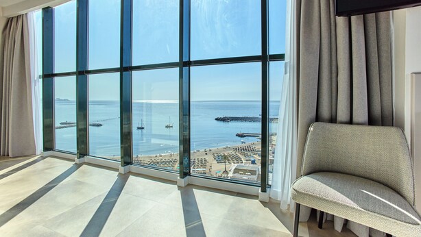 Quarto espaçoso com grandes janelas do chão ao teto, com vista para o mar e um cais.