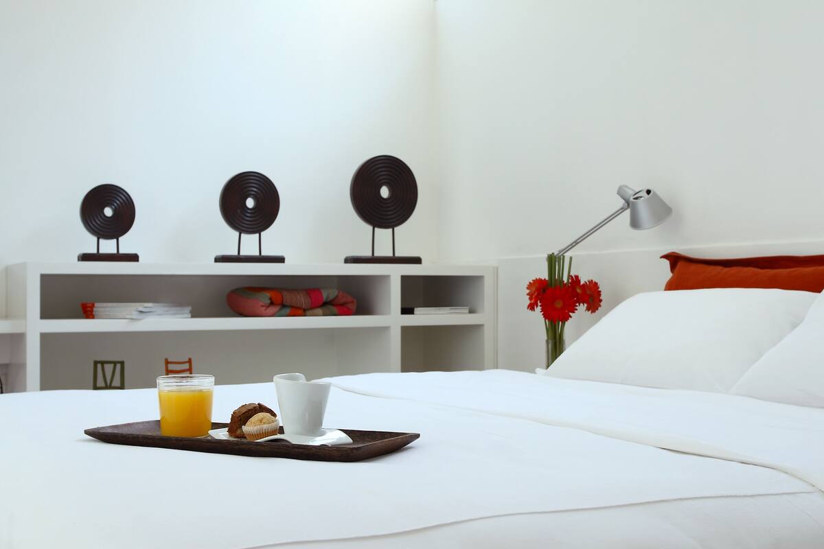 Cama bem arrumada com lençóis brancos, bandeja com itens de café da manhã, mesa de cabeceira com abajur, e parede com objetos decorativos e uma prateleira.