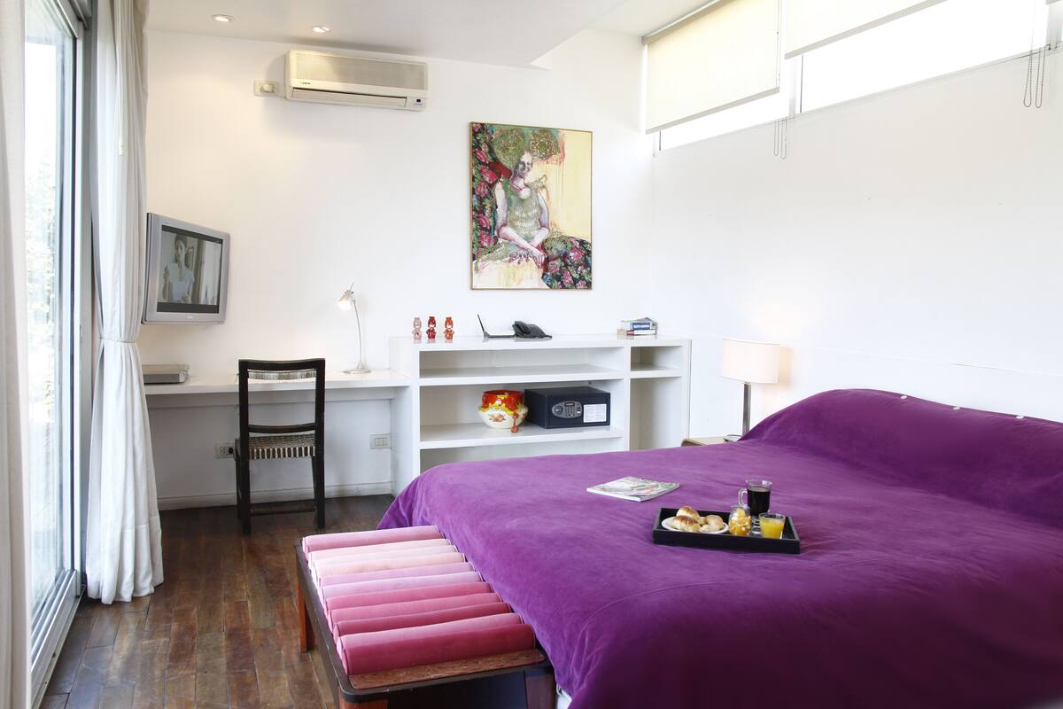 Quarto com colcha roxa, escrivaninha com laptop, cadeira, prateleira com objetos decorativos e janela com cortinas.