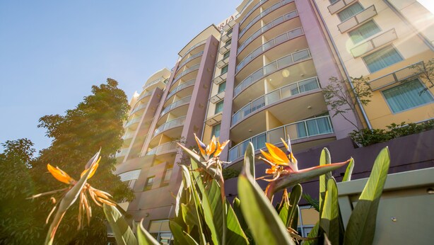 Hotel com vários andares, varandas, céu claro e plantas tropicais em primeiro plano.