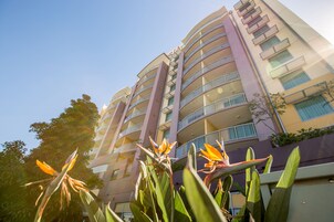 Hotel com vários andares, varandas, céu claro e plantas tropicais em primeiro plano.