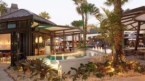 Uma área de refeições moderna ao ar livre com piscina, palmeiras e um espaço coberto para relaxar.