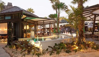 Uma área de refeições moderna ao ar livre com piscina, palmeiras e um espaço coberto para relaxar.