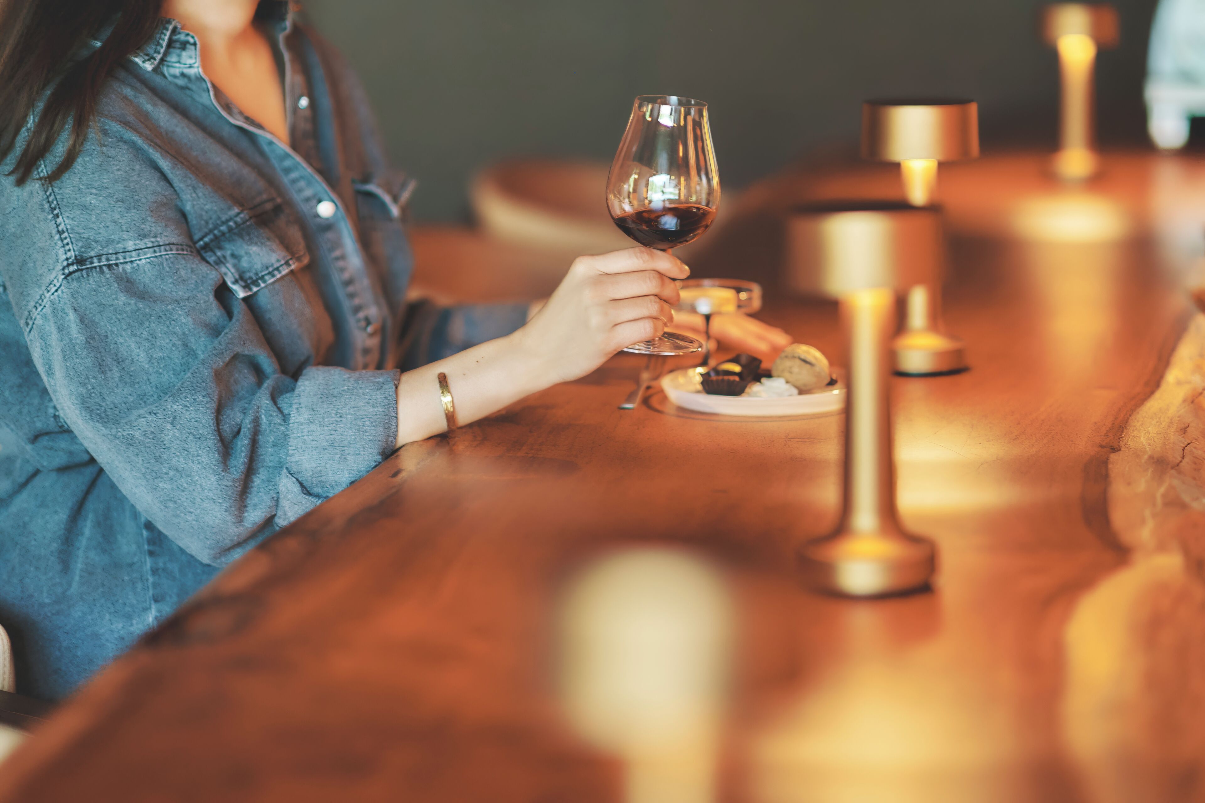 Uma pessoa sentada à mesa de madeira com uma taça de vinho, um prato de comida e um abajur.