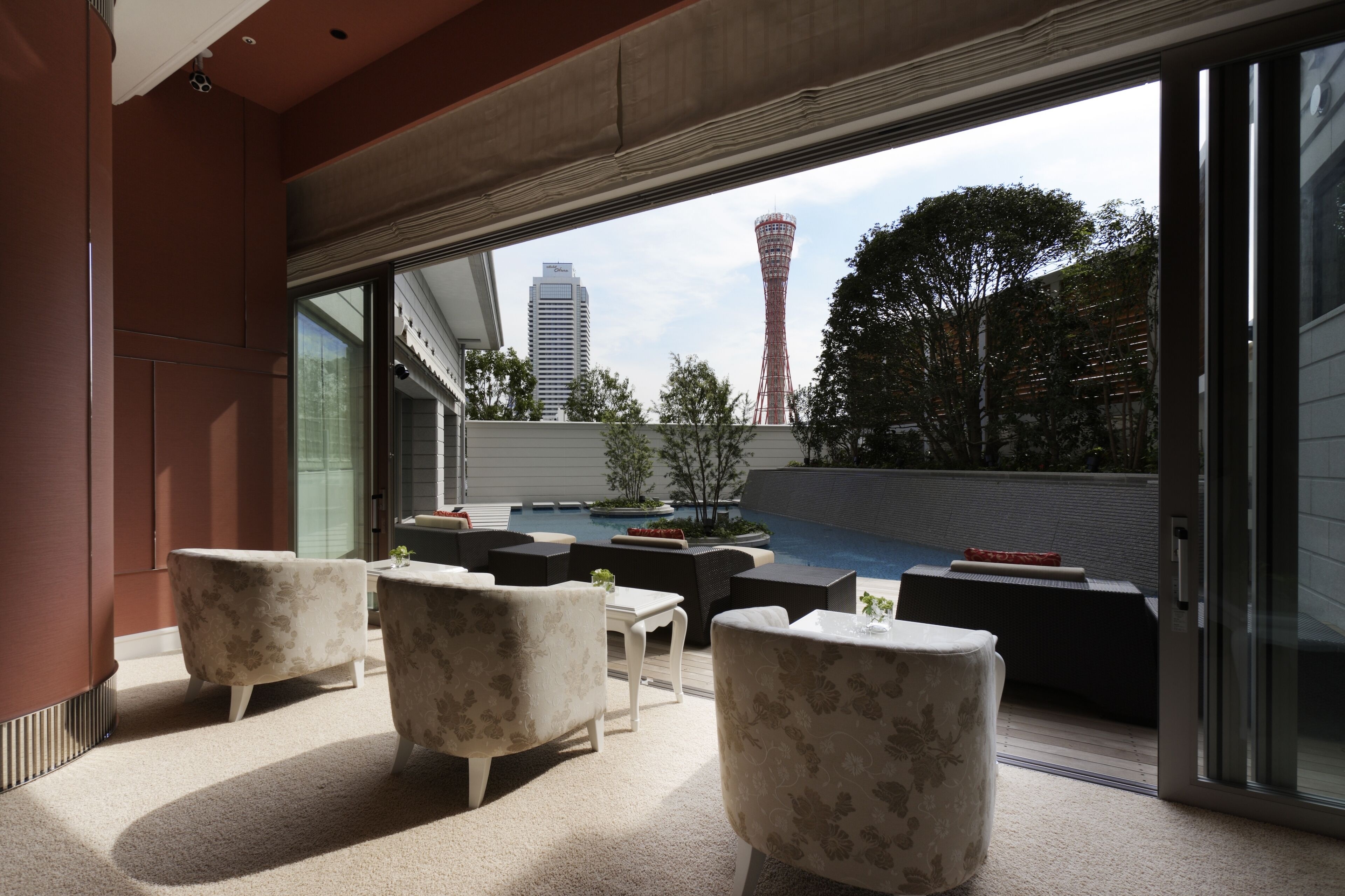 Uma área de estar moderna ao ar livre com sofás e uma pequena mesa, com vista para uma piscina e um horizonte urbano com uma torre alta.