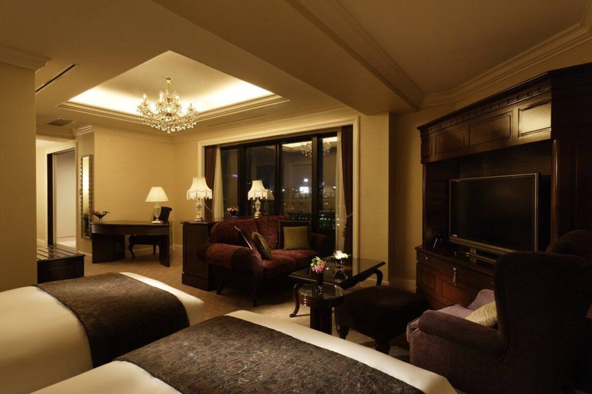 Quarto de hotel com duas camas, televisão, lustro e vista da cidade à noite.