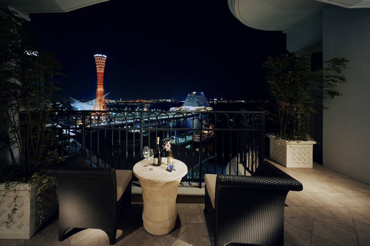 Terraço no telhado com móveis de vime, uma mesa com garrafa e copos, e vista para a cidade à noite.