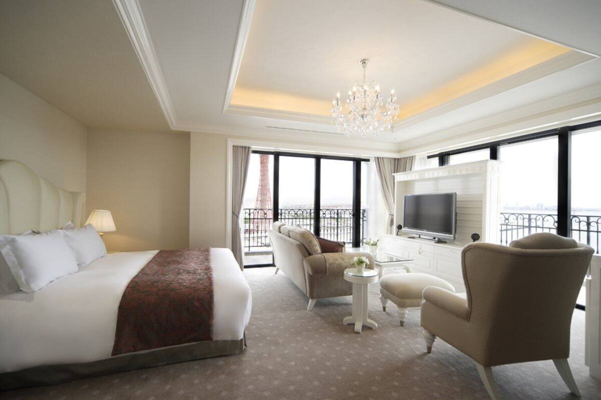Quarto de hotel com uma cama grande, um sofá, uma poltrona, uma mesinha, uma televisão e uma varanda com vista.
