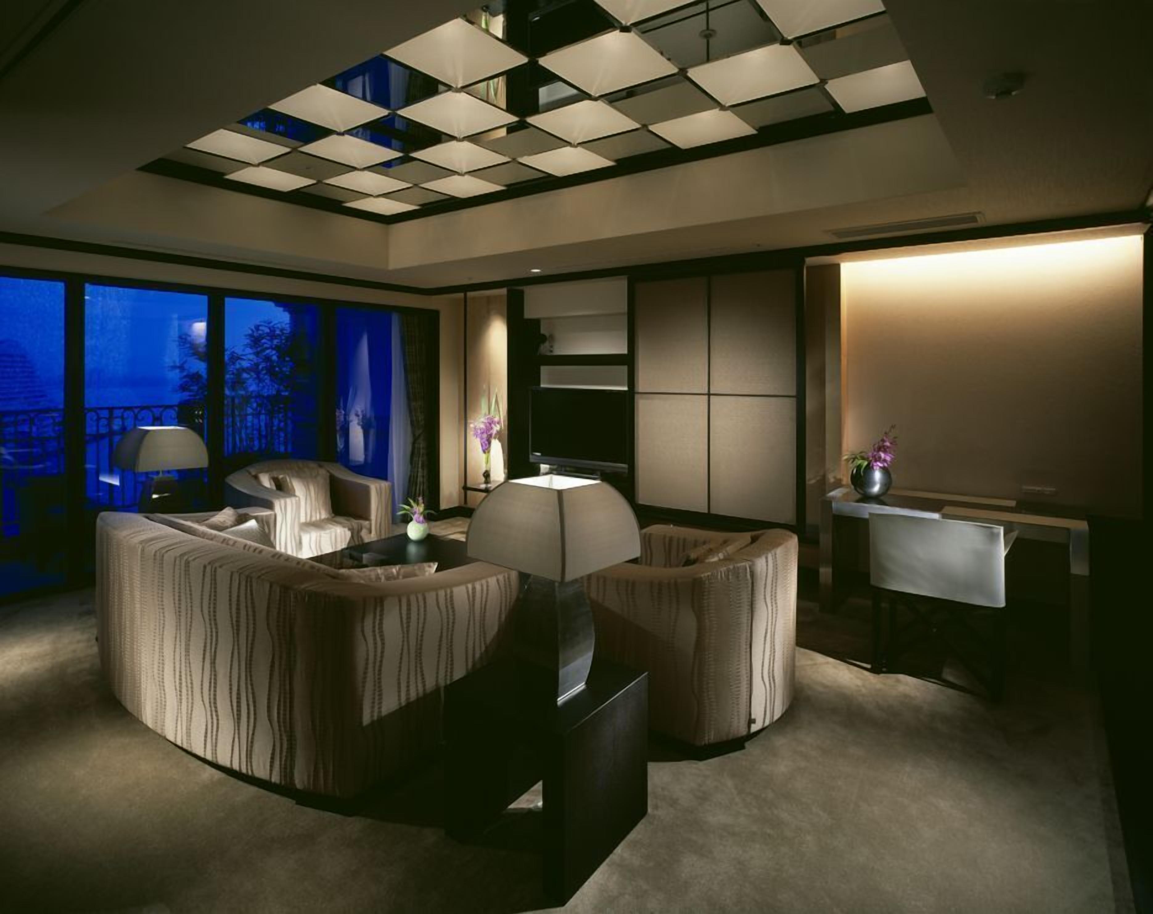 Uma sala de estar moderna com um sofá, poltronas e uma mesa de centro. Há uma televisão e uma pequena mesa com um vaso. O quarto tem uma janela grande com vista para um edifício.